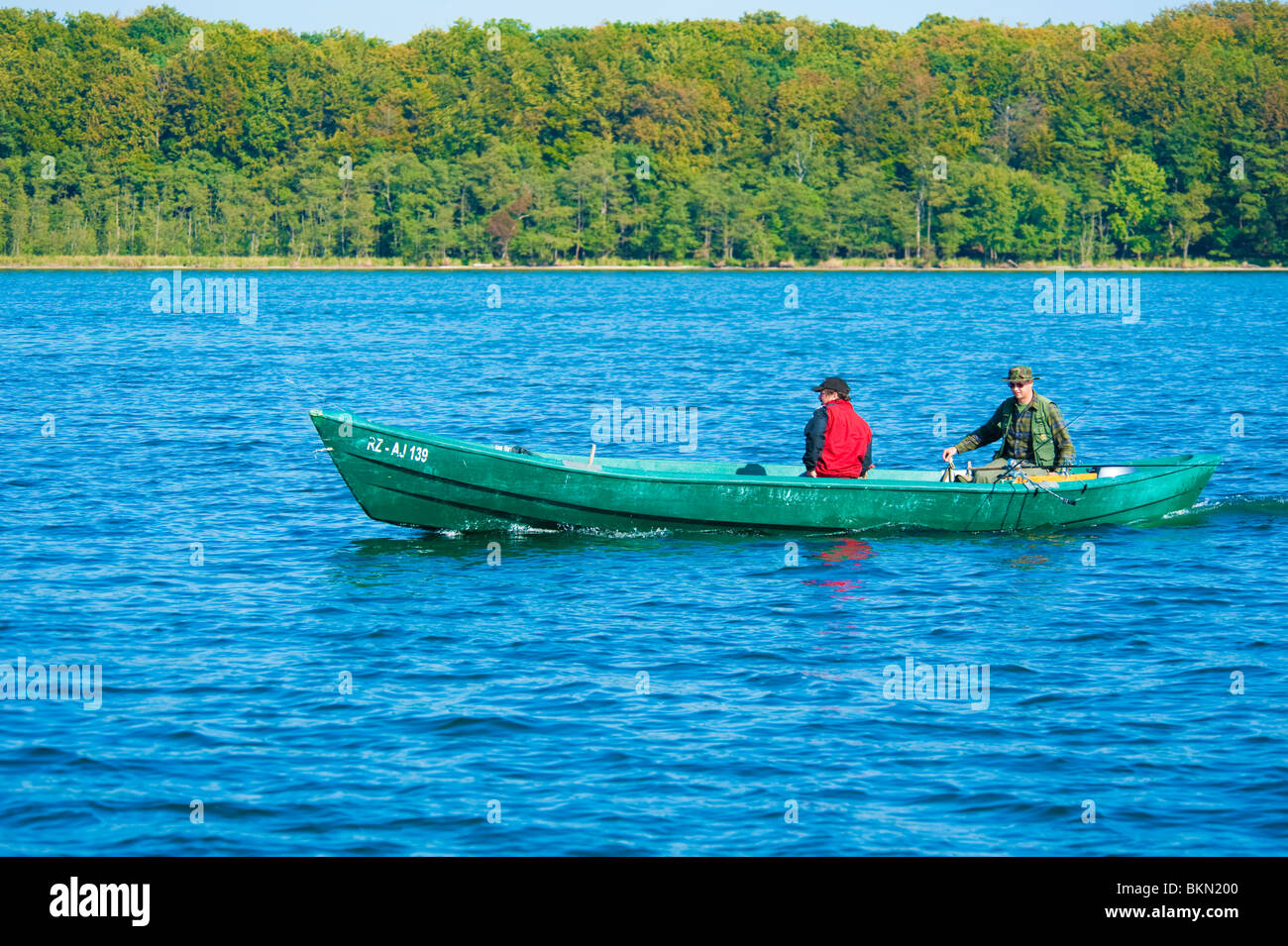 Two fishermen in green fishing boat in front of forrest, lake near ...