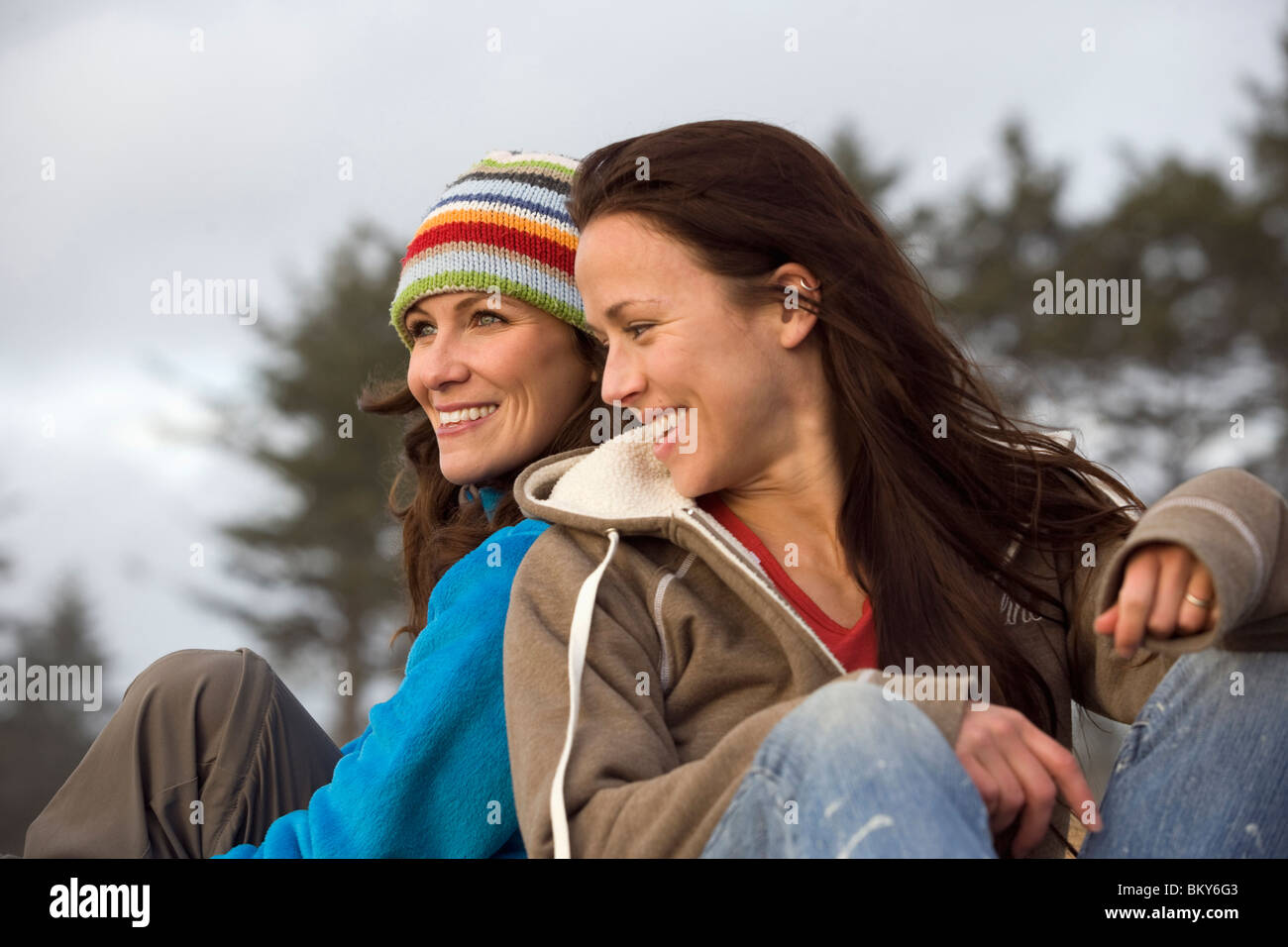 Two young women sit back to back in a open forest setting, smiling and ...