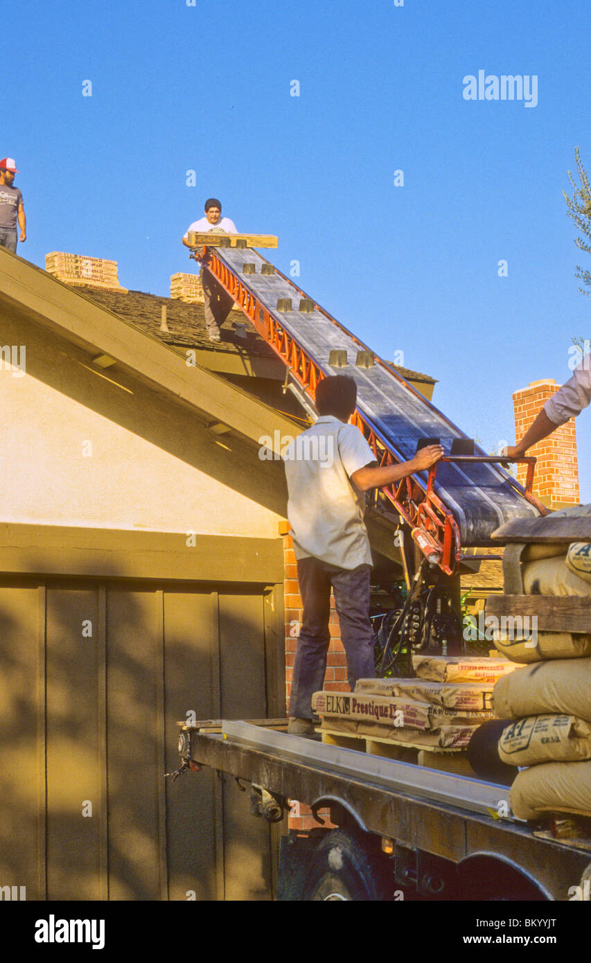 Hispanic workers use conveyor belt to place roofing shingles on roof of ...