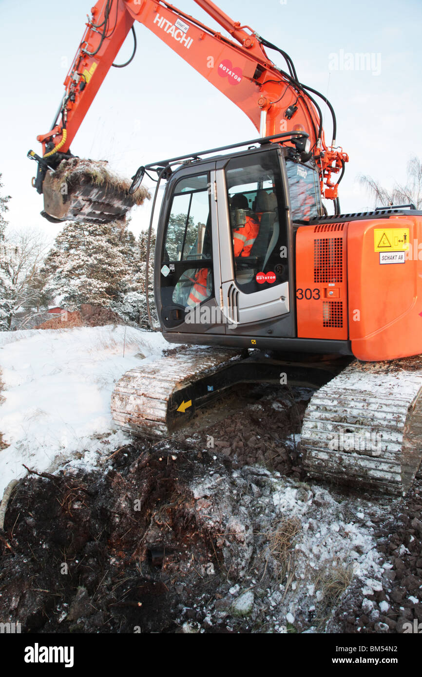 A digger excavator digs a trench in the earth in heavy winter snow ...