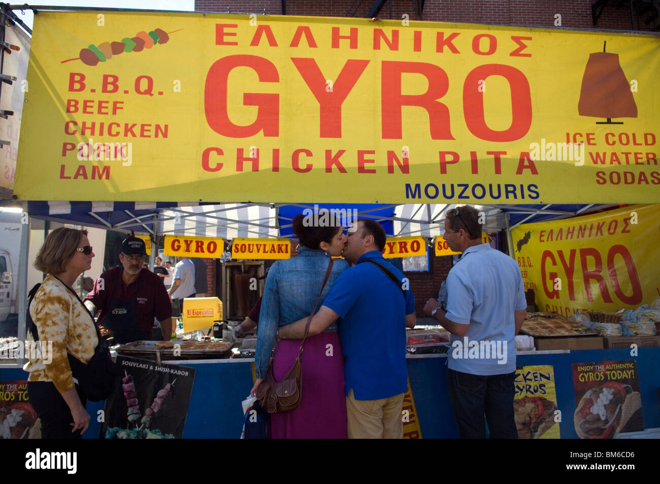 A couple kisses in front of a stand selling Greek food, gyros, and pita ...