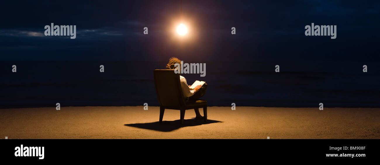 Man sitting in chair under light bulb on beach at night, reading book ...
