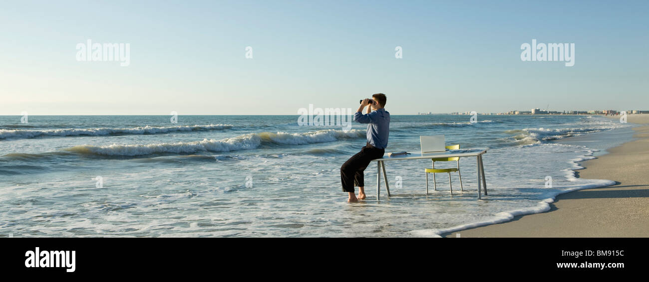 Businessman sitting on desk at the beach, looking through binoculars at ...