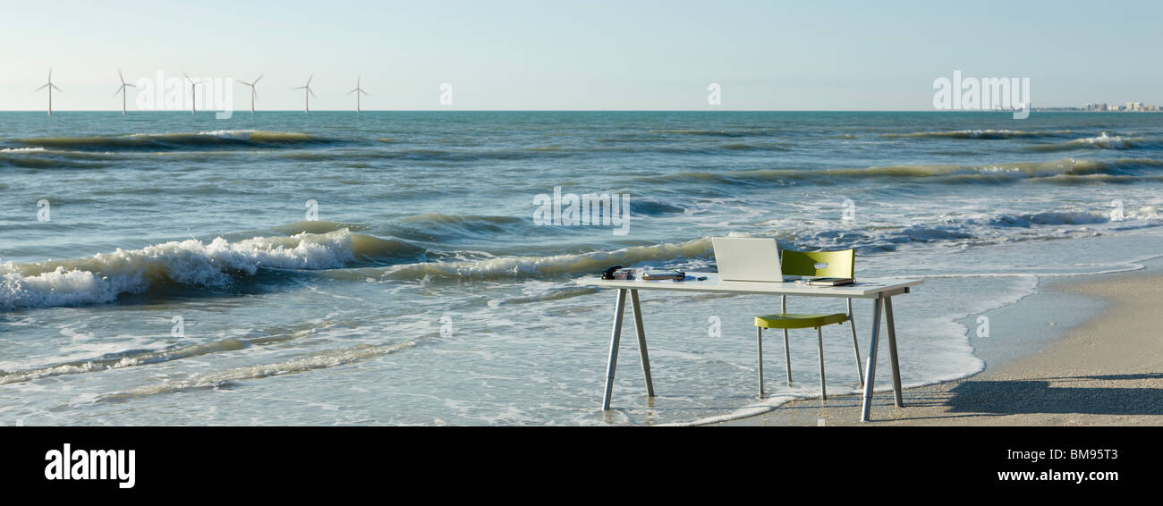 Desk and chair with laptop computer set at water's edge on beach, wind ...
