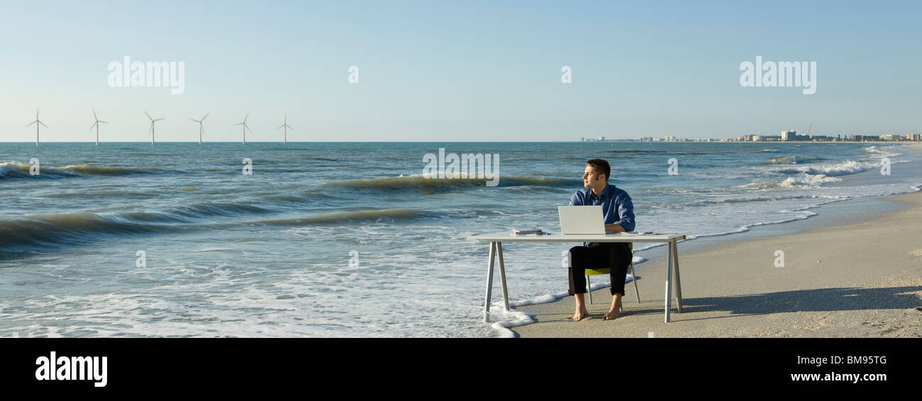 Man working at desk placed at water's edge on beach, wind turbines on ...