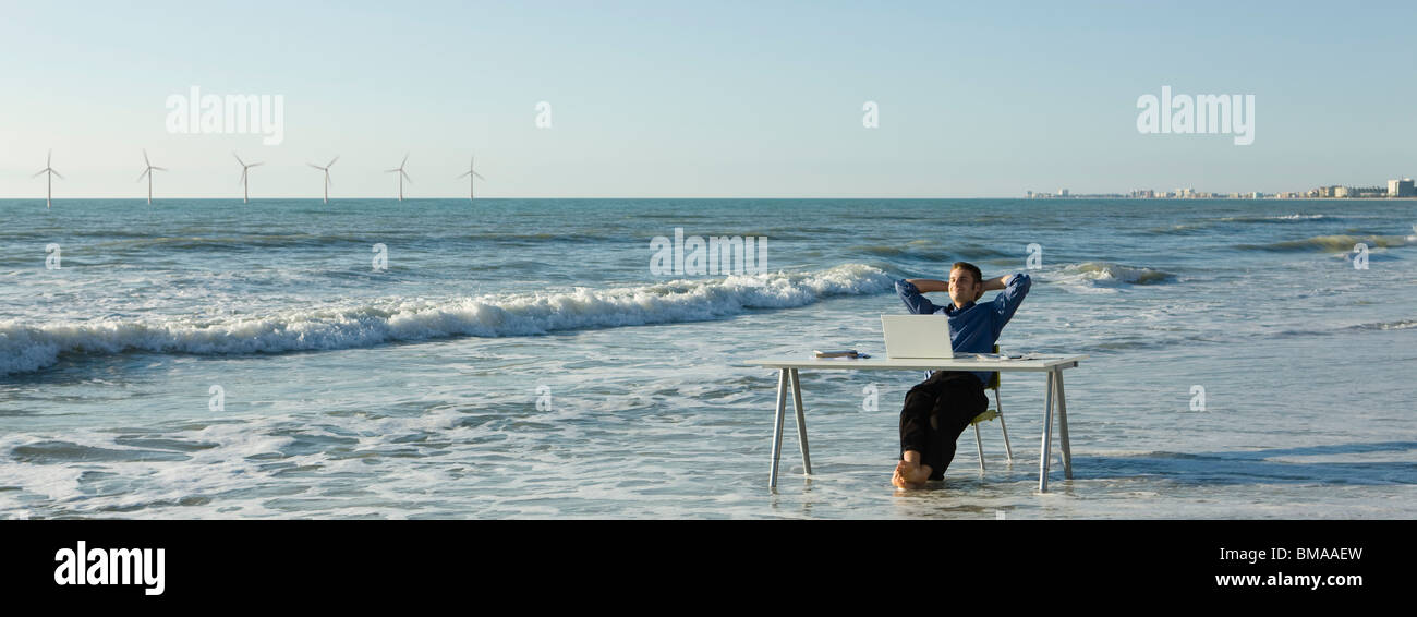 Man relaxing at desk set at water's edge on beach, wind turbines on ...