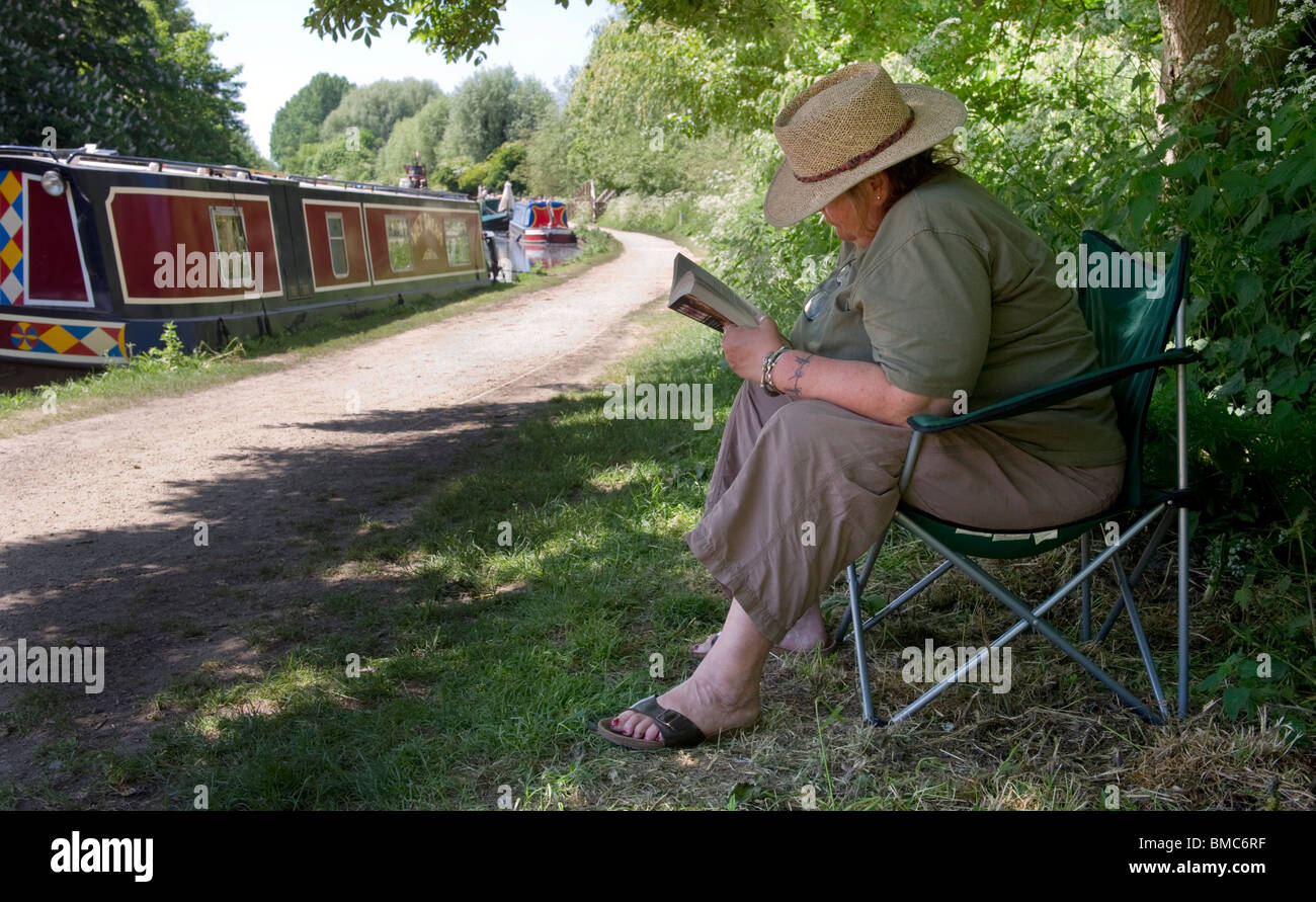 A woman sitting, relaxing and reading a book by the Grand Union canal ...
