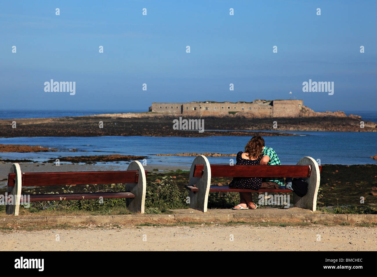 Young couple sitting on the bench in Longis Bay overlooking Ile de Raz ...