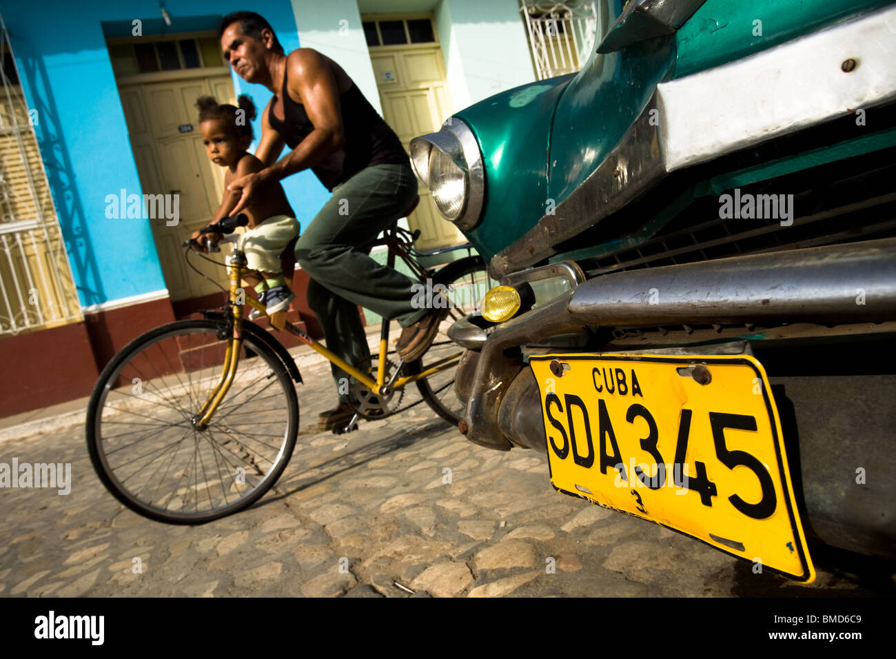 A man and a young girl ride a bicycle past a 1950s automobile parked on ...