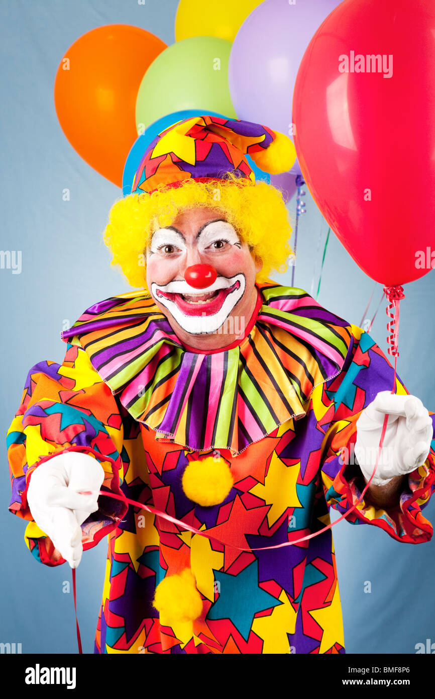 Clown with a bunch of helium balloons, holding a red one toward the ...
