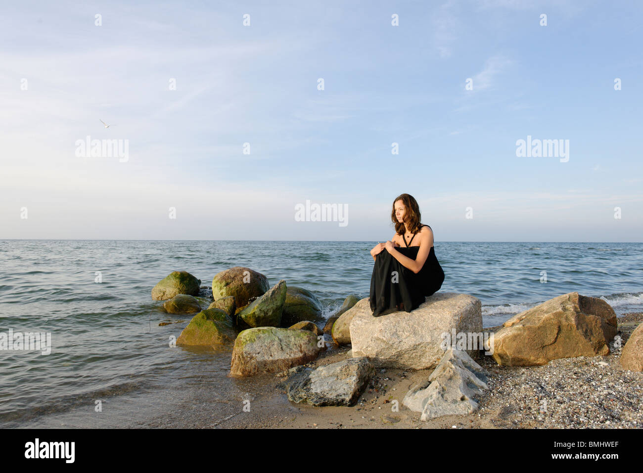 Young woman, 20+, crouching on a rock on a beach, wearing a black dress ...