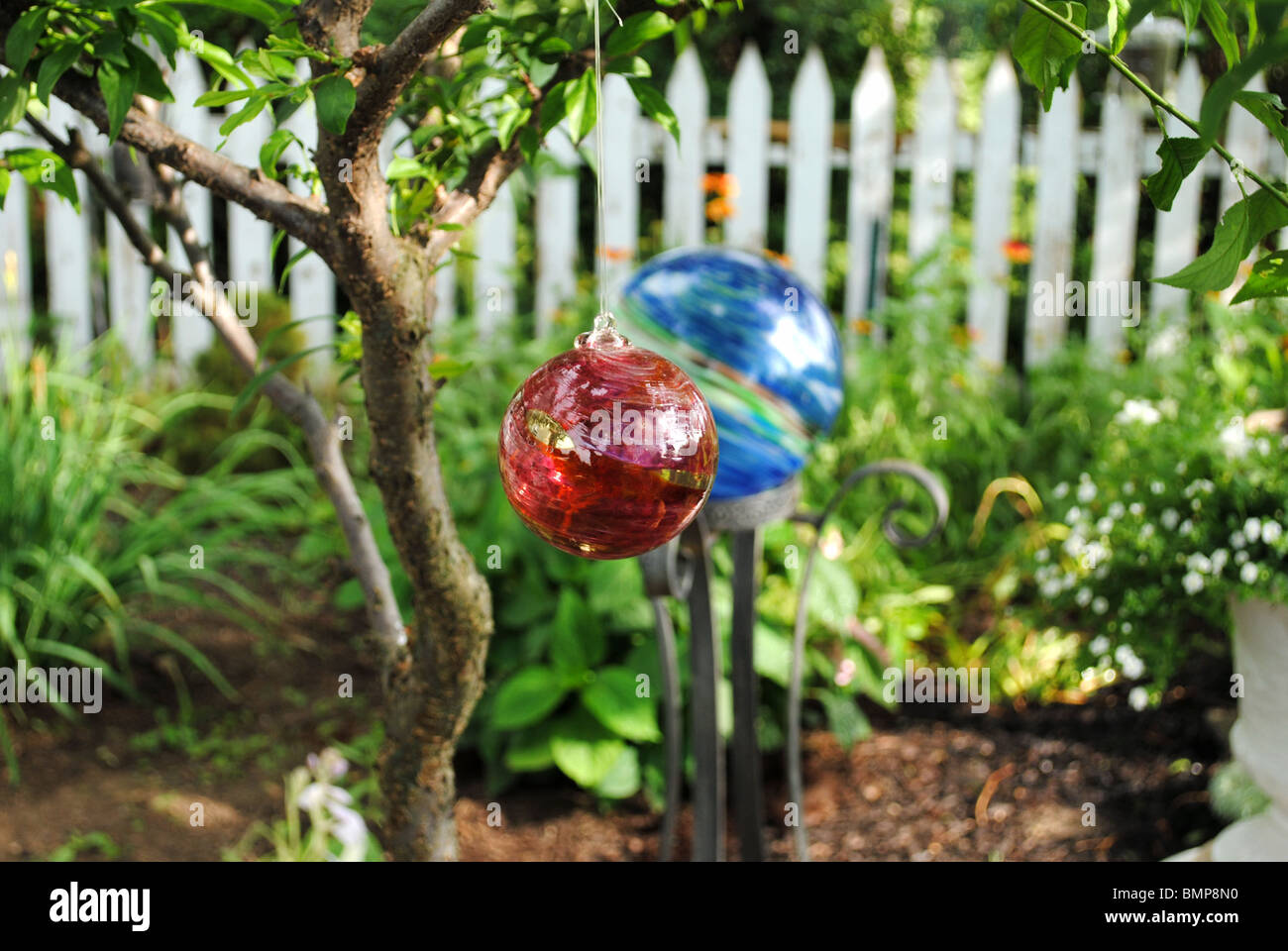 Red hanging glass ball in garden with tree, plants and flowers, picket ...