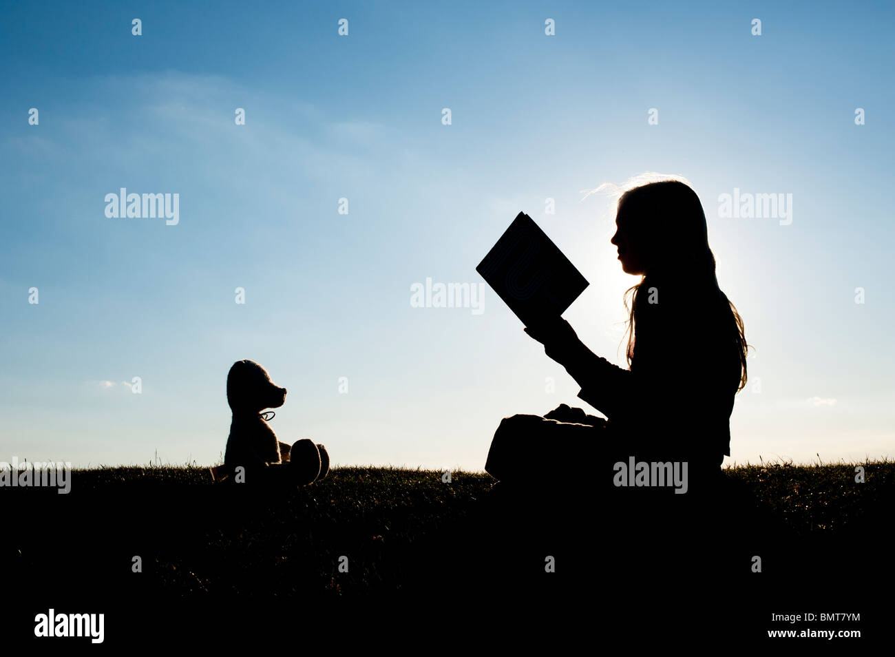 Young Girl sitting down reading a book opposite a teddy bear at sunset ...