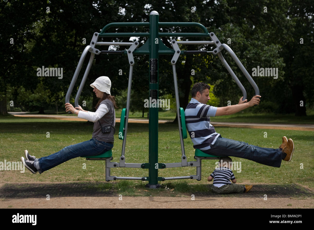 Two parents try out exercise machines in a public park as toddler plays ...