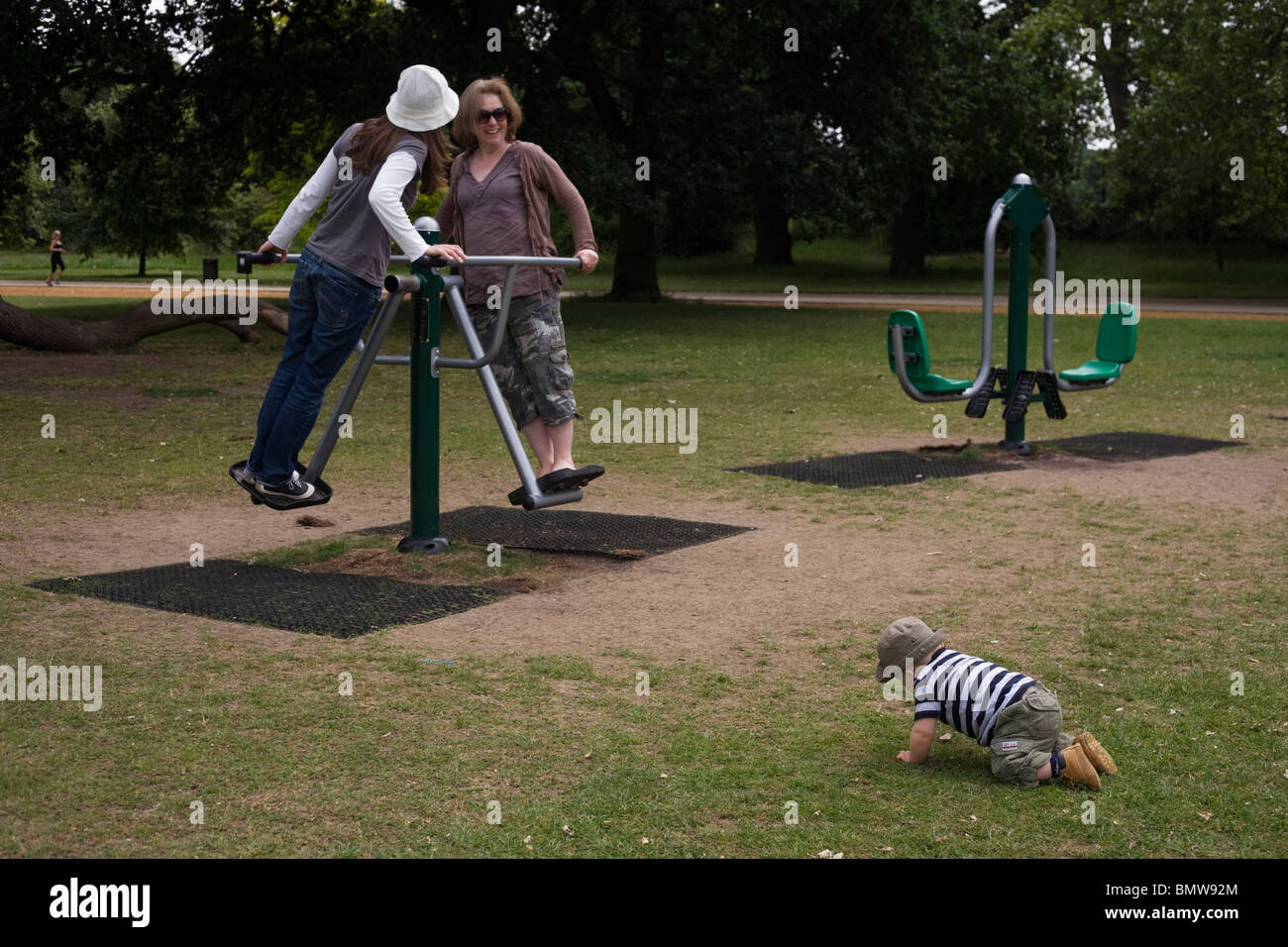Two sisters try out exercise machines is a public park as toddler plays ...