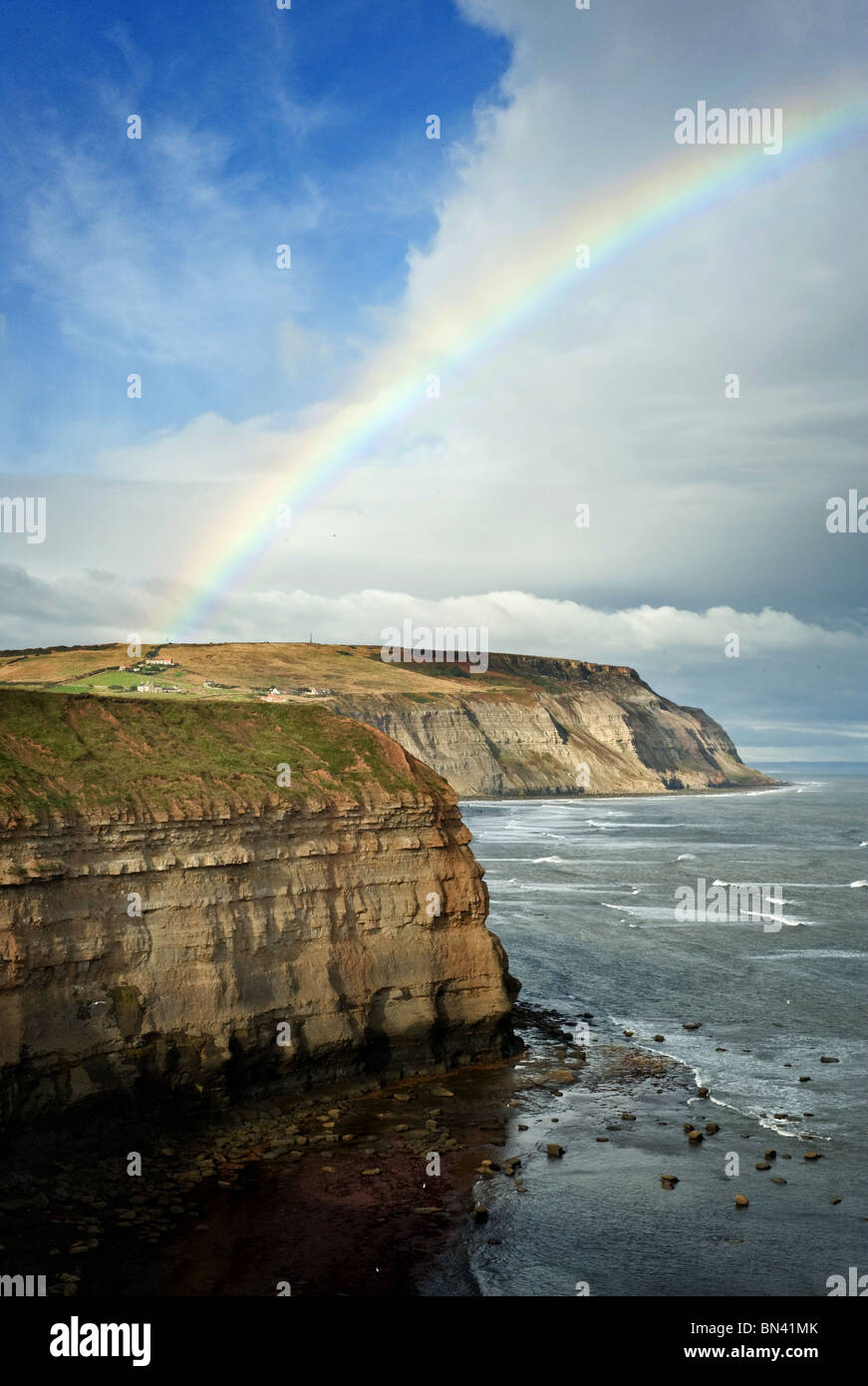 A rainbow reaching out to sea and a cloudy blue sky from a cliff top in ...