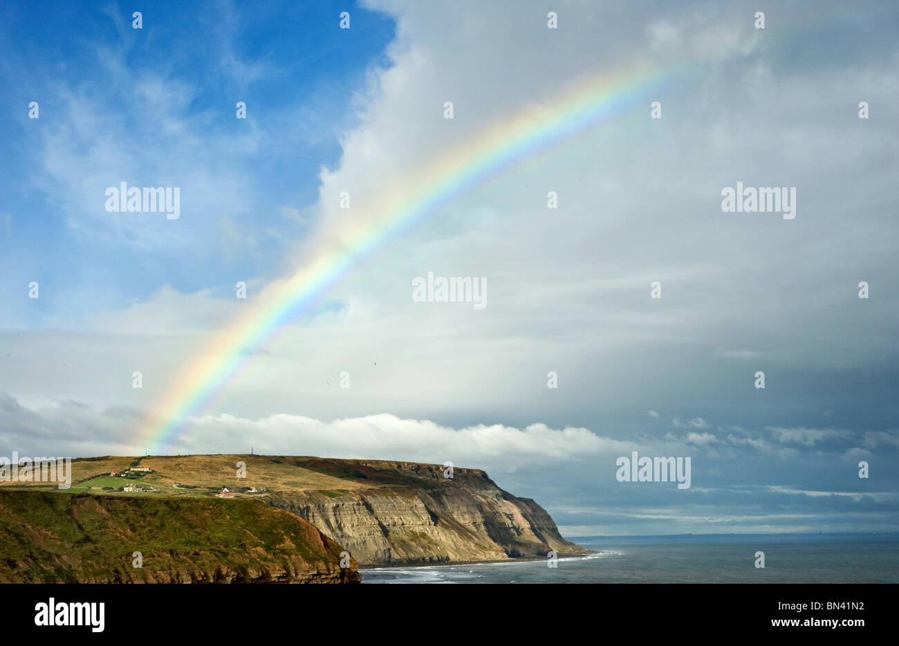 A rainbow reaching out to sea and a cloudy blue sky from a cliff top in ...