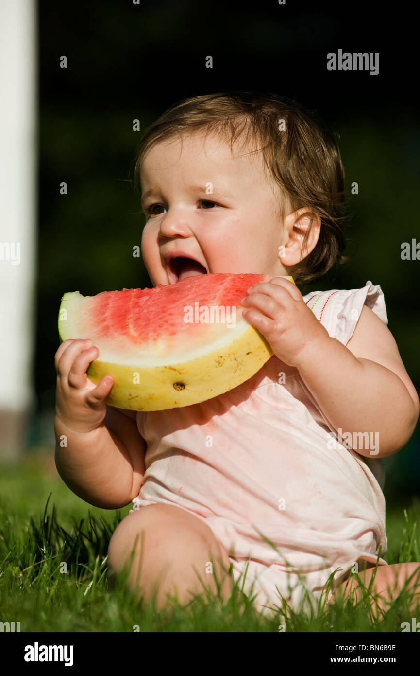 A cute baby, little girl, sits in a grassy field eating watermelon on a ...
