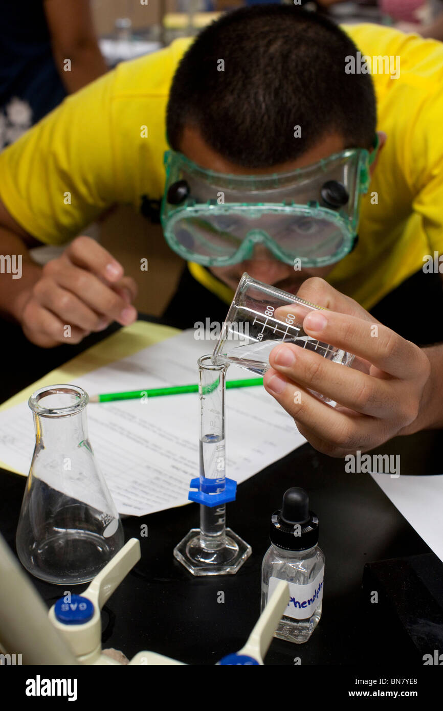 Hispanic male student wearing safety goggles pours solution from beaker ...