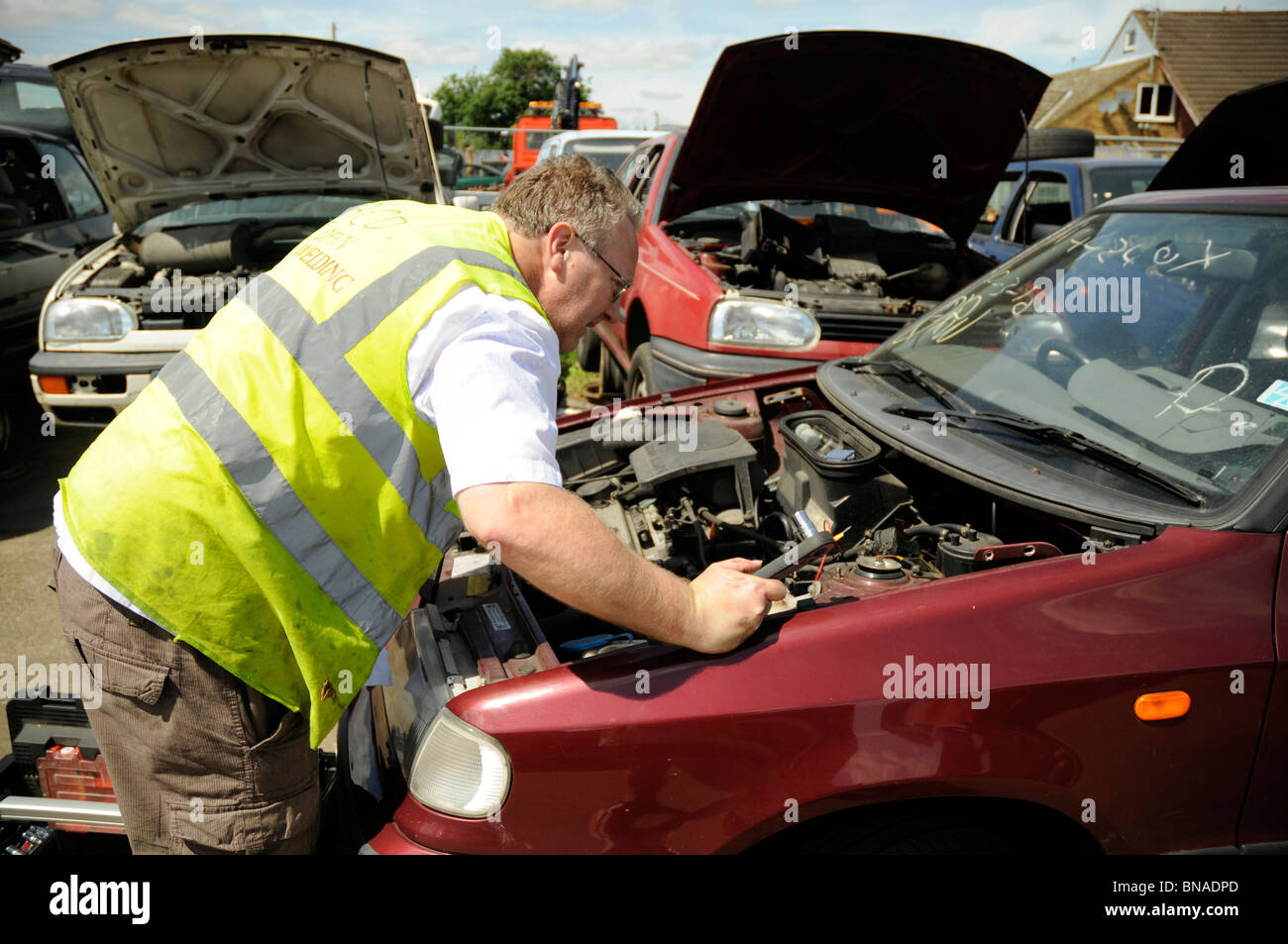 Man wearing hi-vis vest inspecting engine in a car in breakers yard ...