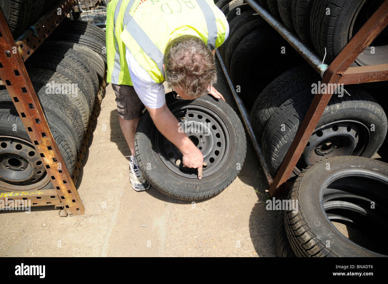 Man wearing hi-vis vest inspecting wheel and tyre in a car breakers ...