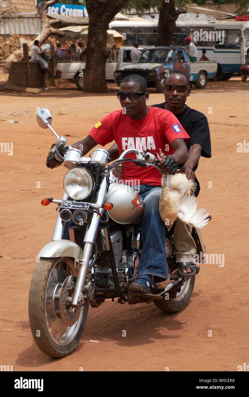 Two African men carrying a chicken while riding motorcycle along a dirt ...