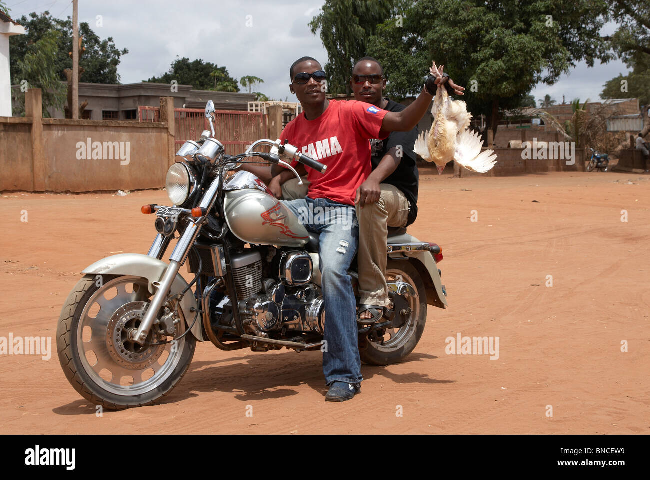 Two African men posing on a motorcycle, driver holding a chicken in an ...