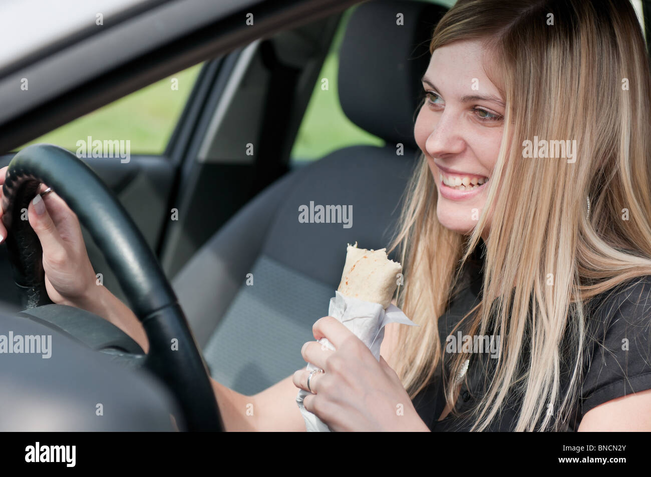 Young beautiful smiling woman driving car and eating fast food ...