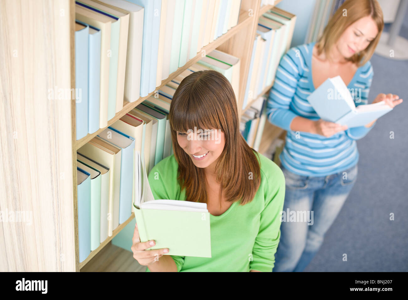 Student in library - two happy woman read book from bookshelf Stock ...
