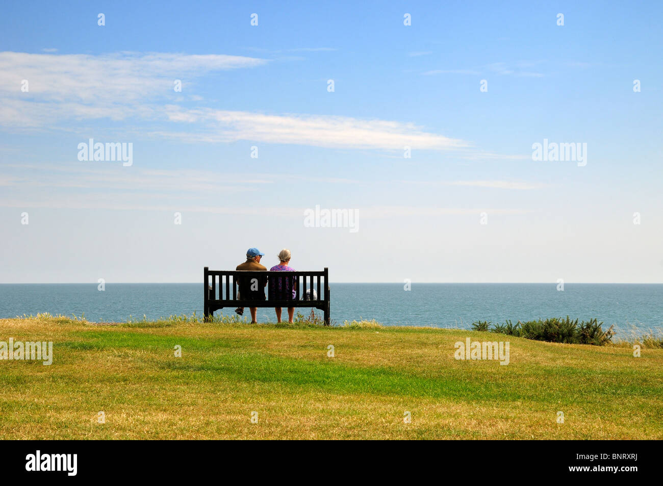 Back view of elderly couple sitting on cliff bench looking out to sea ...