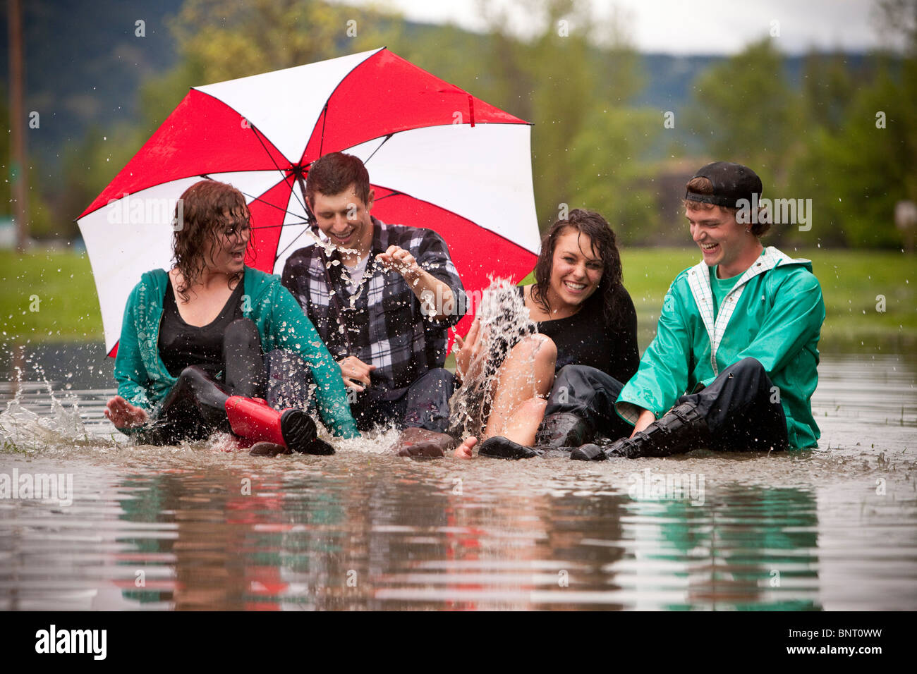 Four young adults laugh while sitting in a large puddle after a water ...