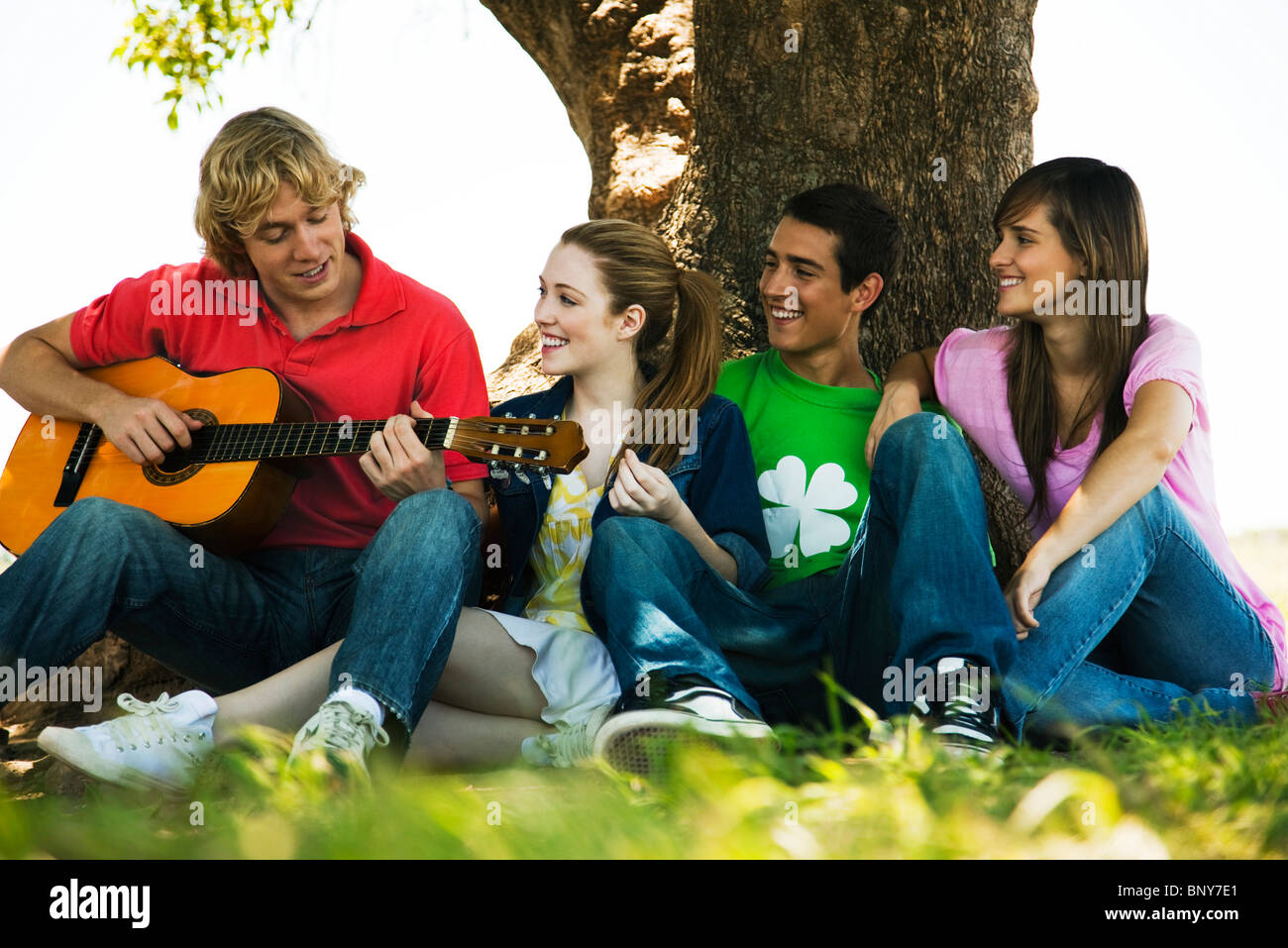 Young man sitting with friends under tree, playing guitar Stock Photo ...