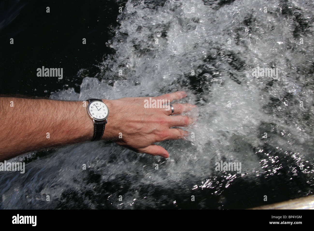 A man's hand draped over the side of a boat letting the water run ...
