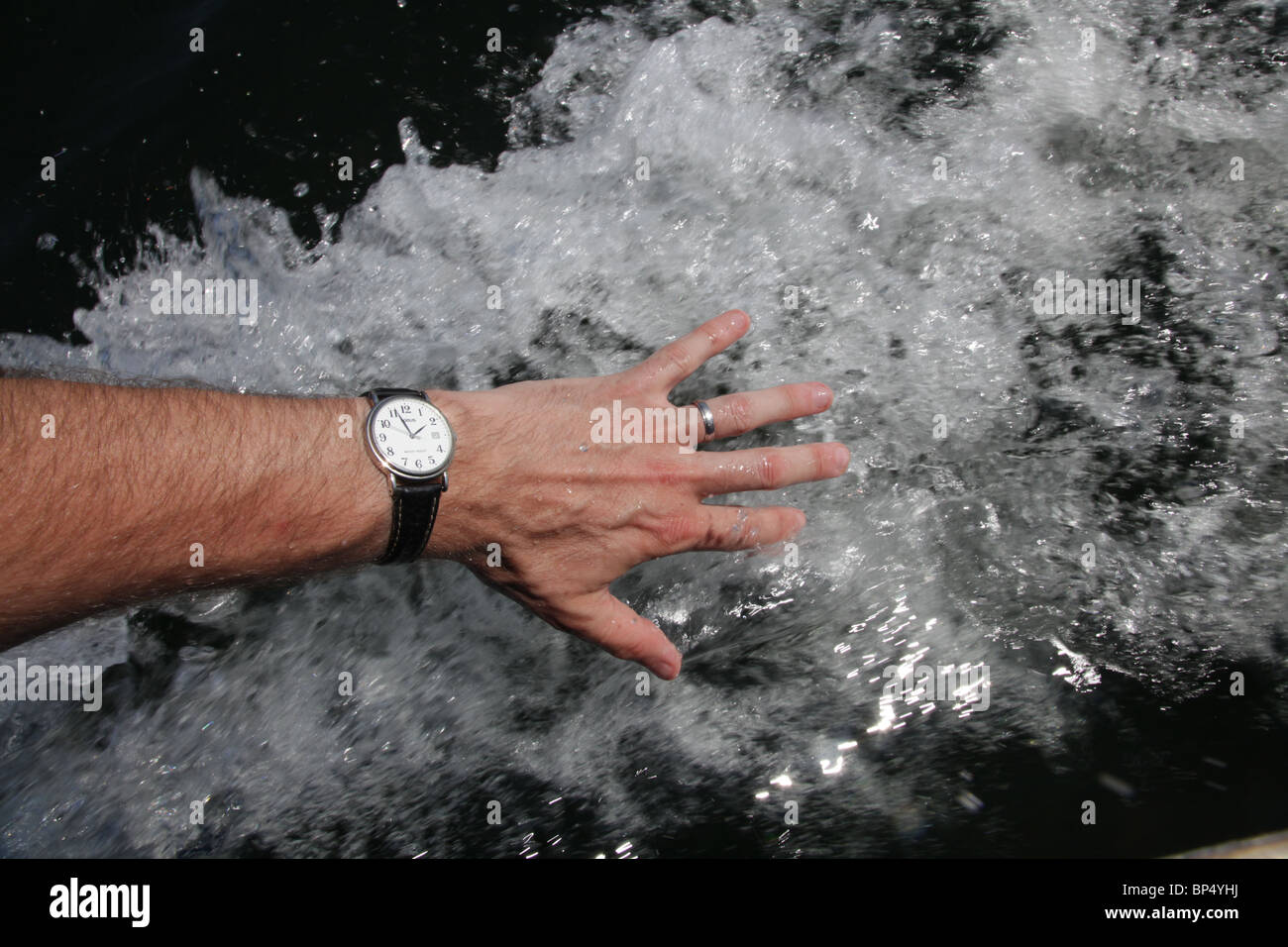 A man's hand draped over the side of a boat letting the water run ...