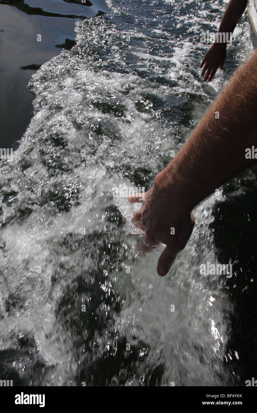 A man's hand draped over the side of a boat letting the water run ...