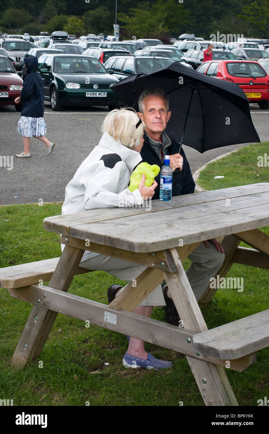 Man and woman sitting on a park bench next to a car park in the rain ...