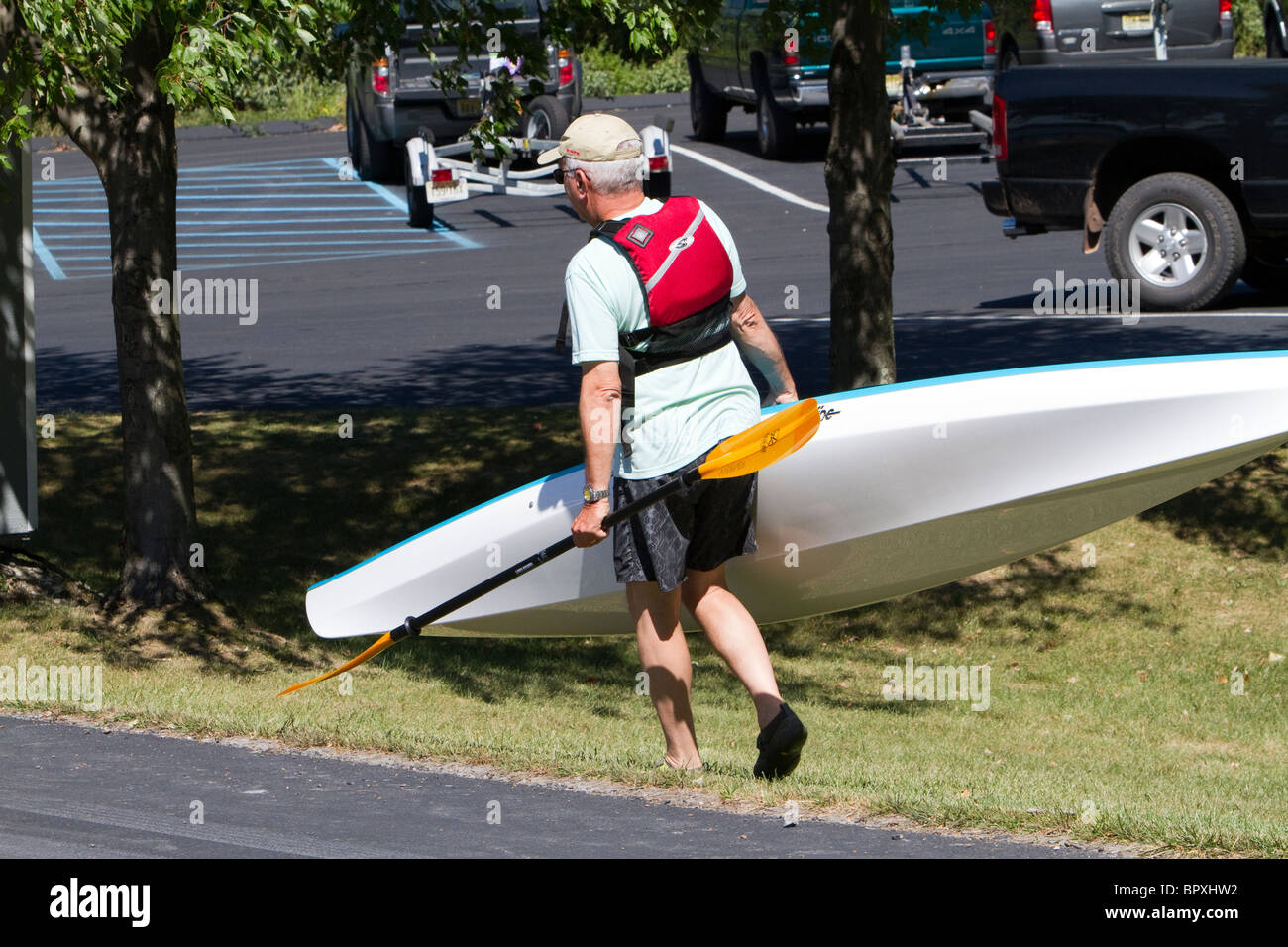 A senior man carrying white kayak and paddles on the side of the road ...