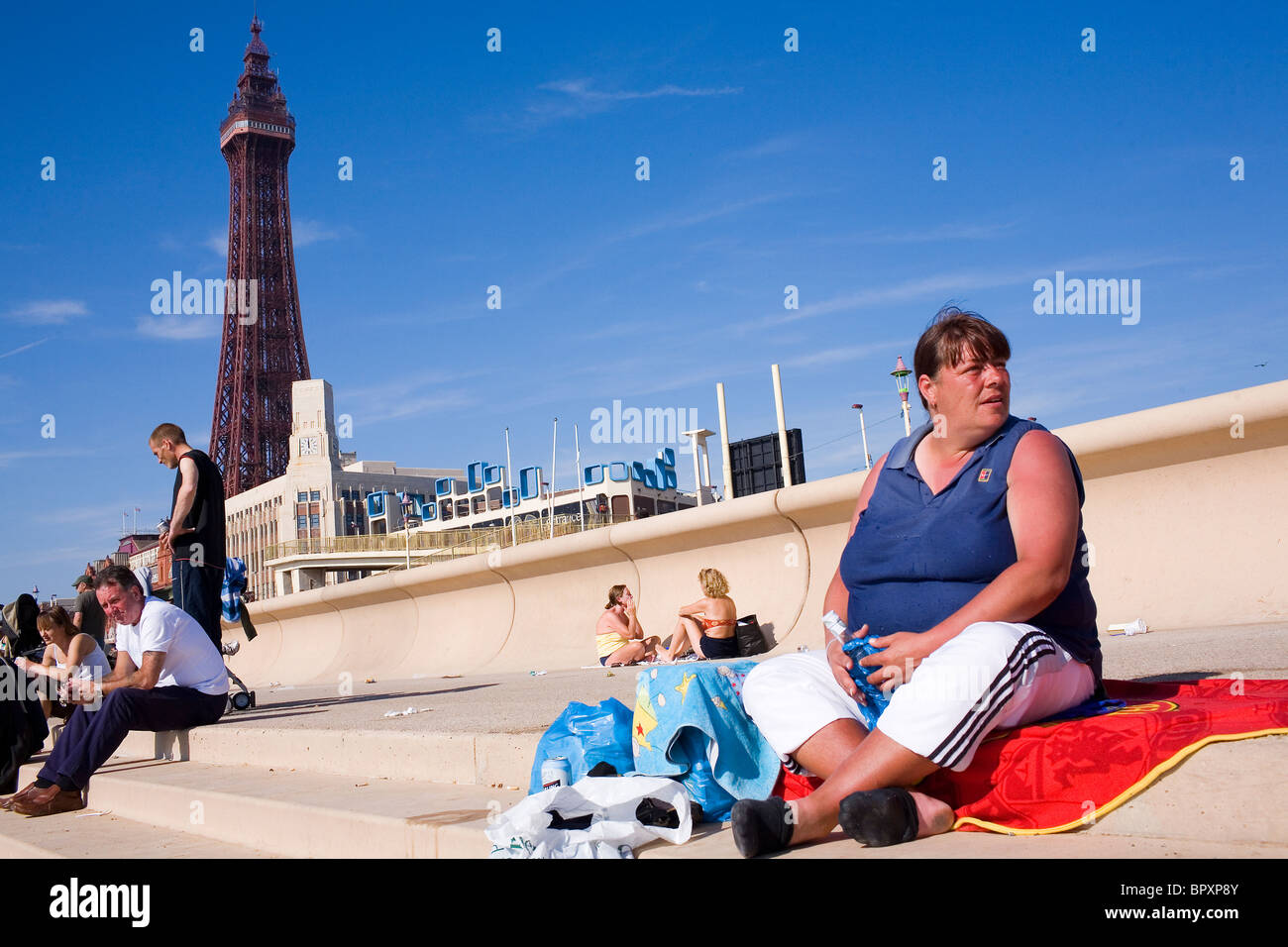 A large woman sits on a red towel on the promenade near Blackpool Tower ...