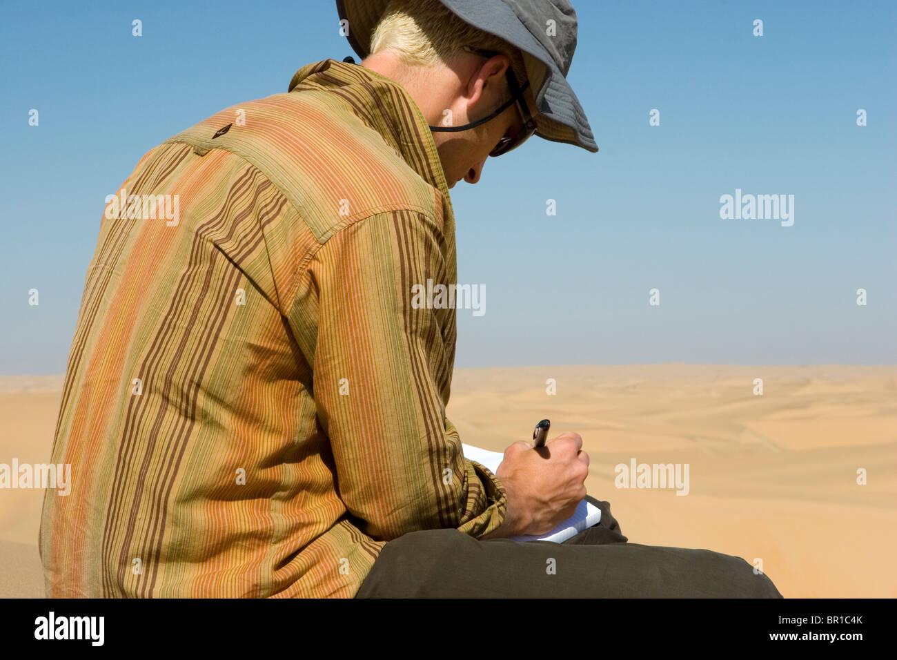 A man sits atop the dunes on the Namib Desert, Namibia writing in his ...