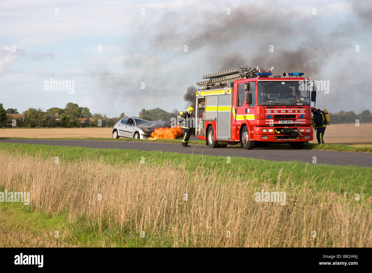 Car on roadside verge with it's engine on fire. Fire Brigade having ...