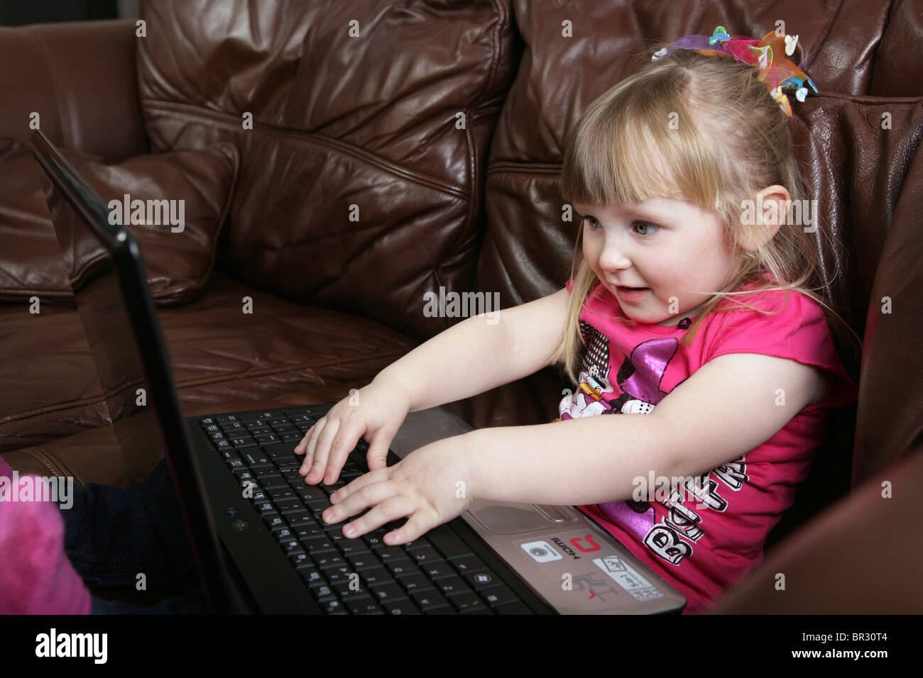 A three year old girl sitting on a sofa with a PC laptop on her lap ...