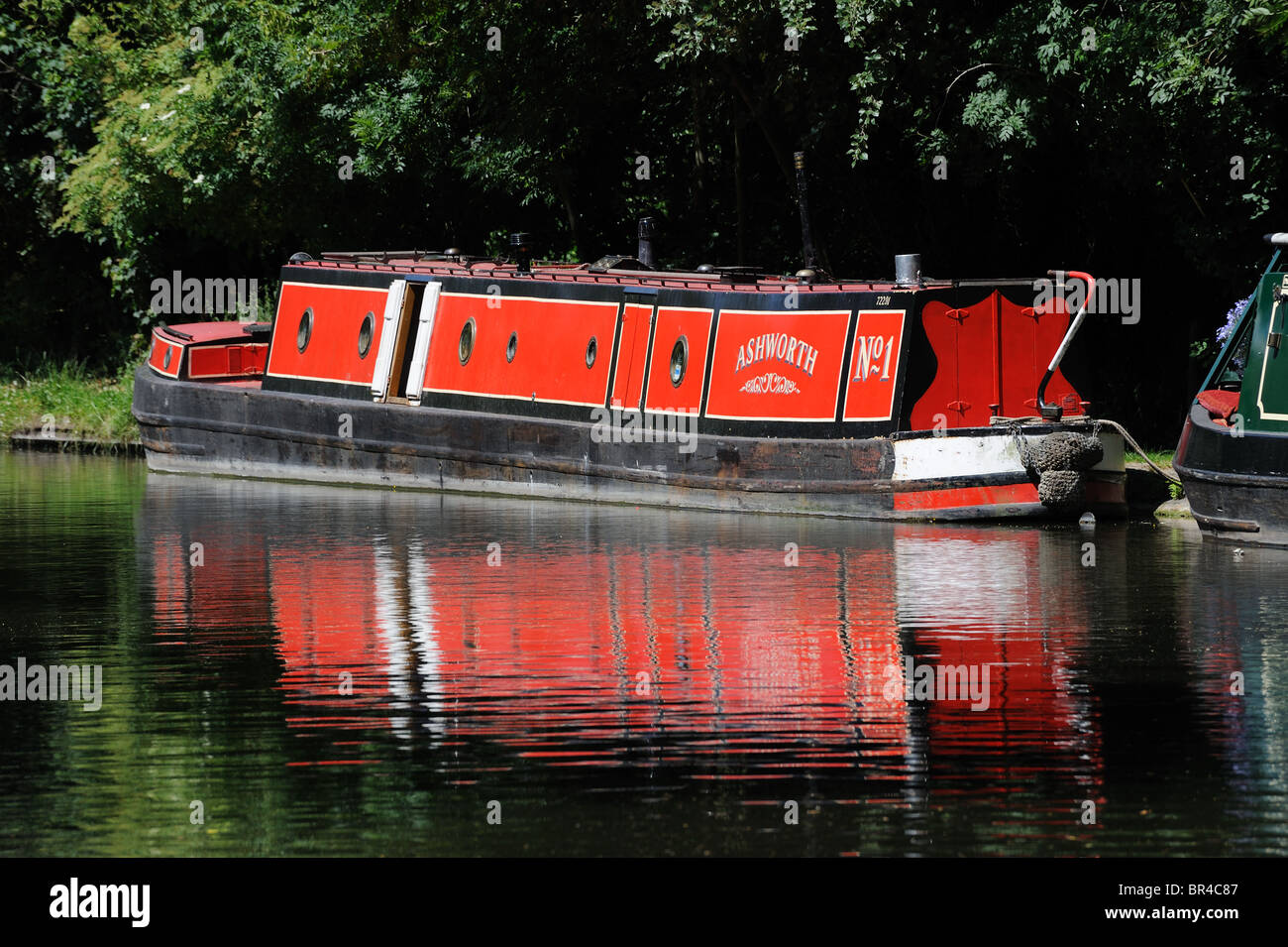 A bright red narrowboat moored below trees on the Grand Union Canal ...