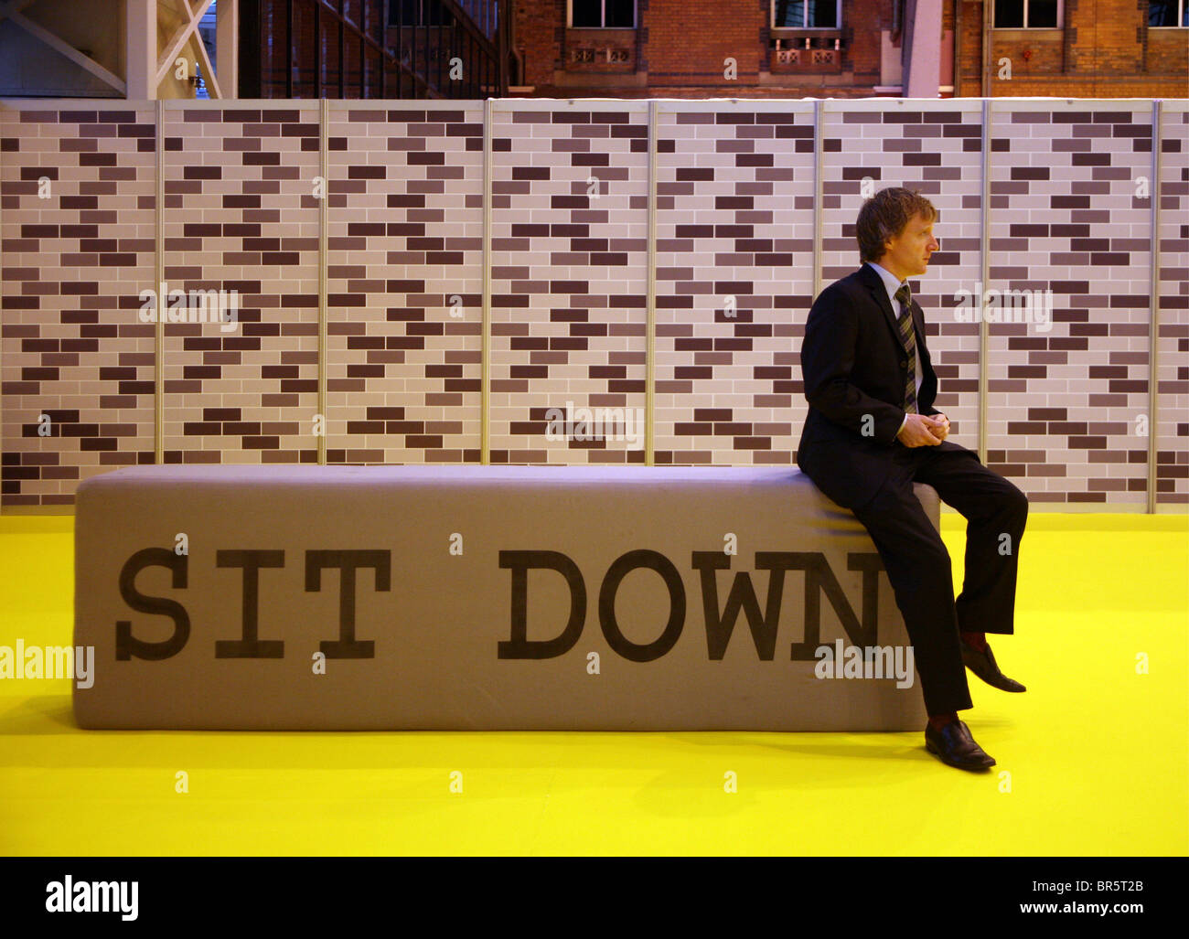A man sits down on a 'Sit down’ sign located at the entrance of a ...