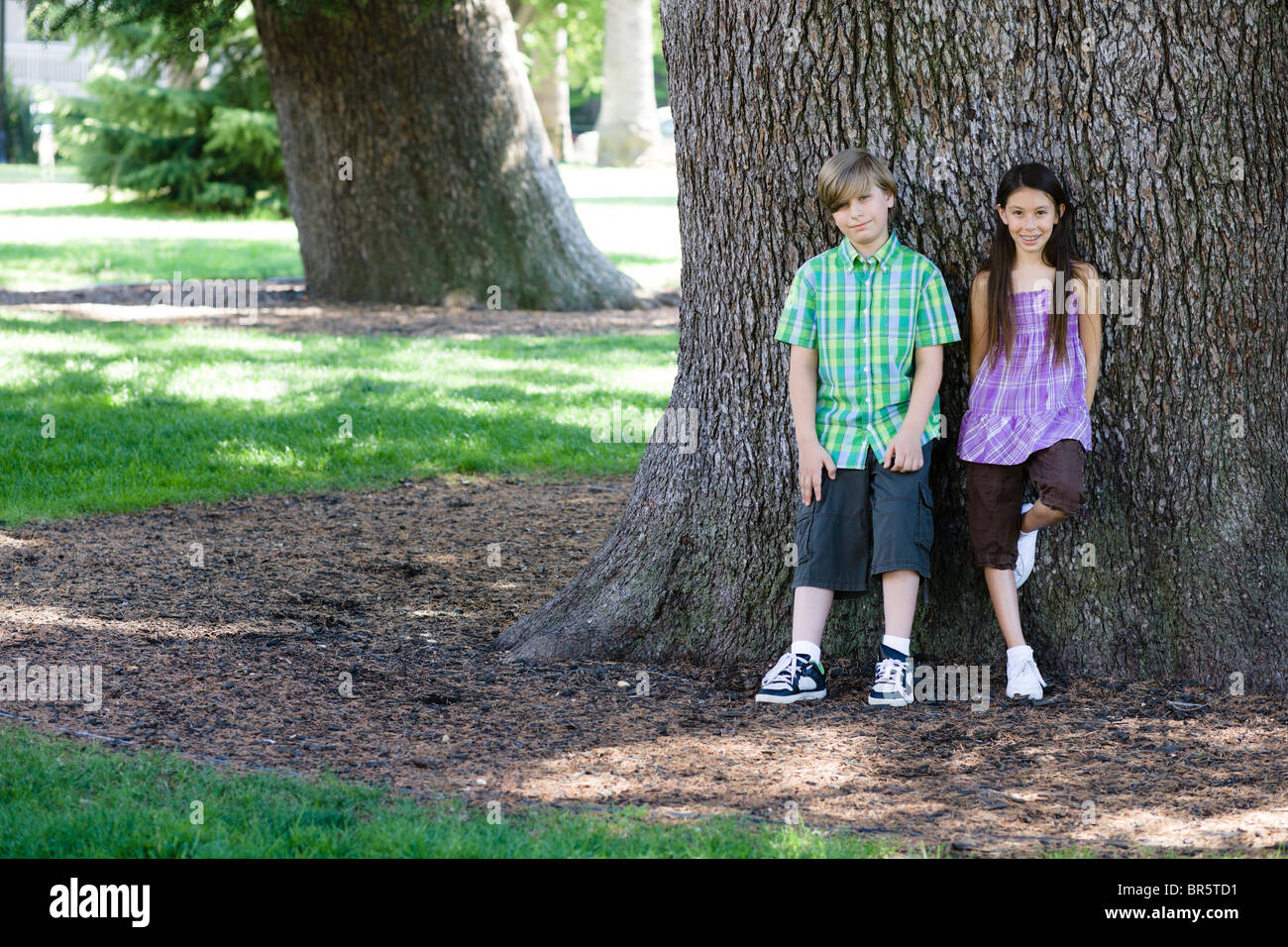Boy and girl leaning against tree trunk in park Stock Photo - Alamy