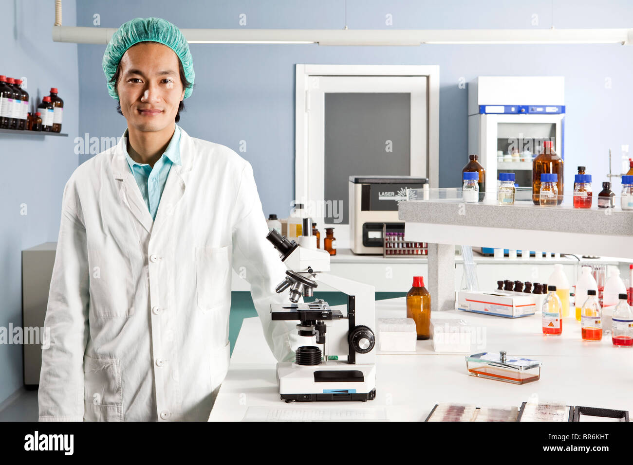 A lab technician standing next to a microscope, looking at camera Stock ...