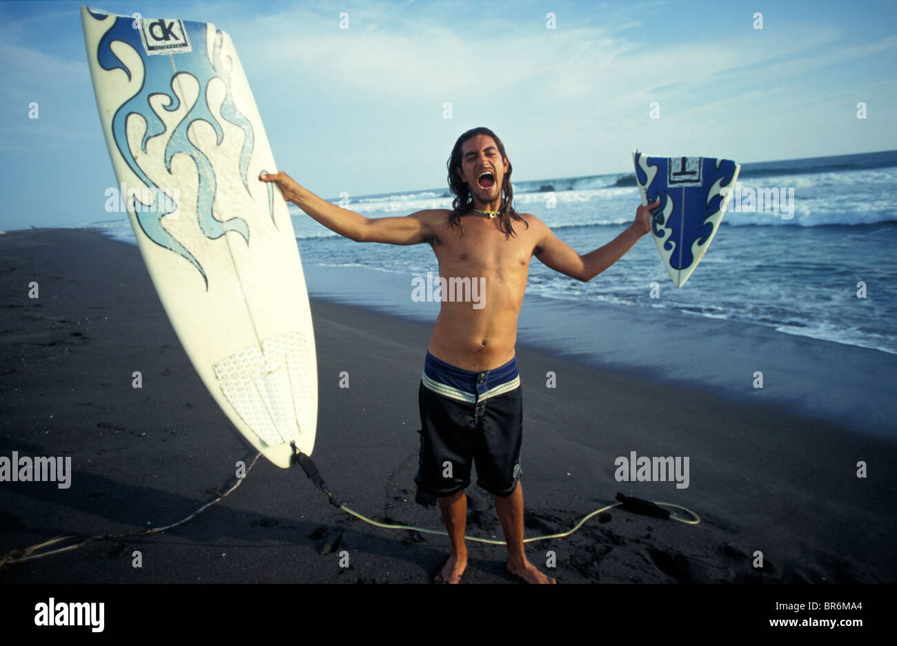 A male surfer screams and holds up his broken surfboard on the beach ...