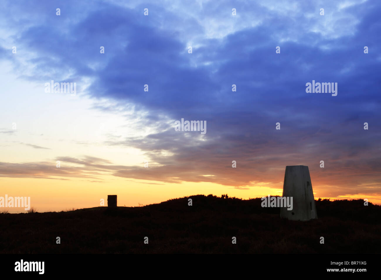 Trig point and commemorative stone in silhouette against colourful sky ...