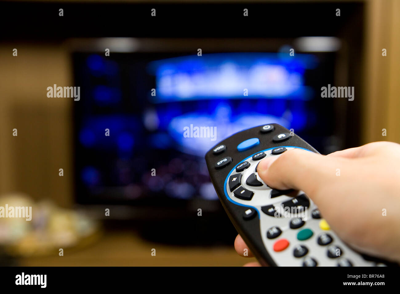 Hand holding TV remote control with a television in the background ...