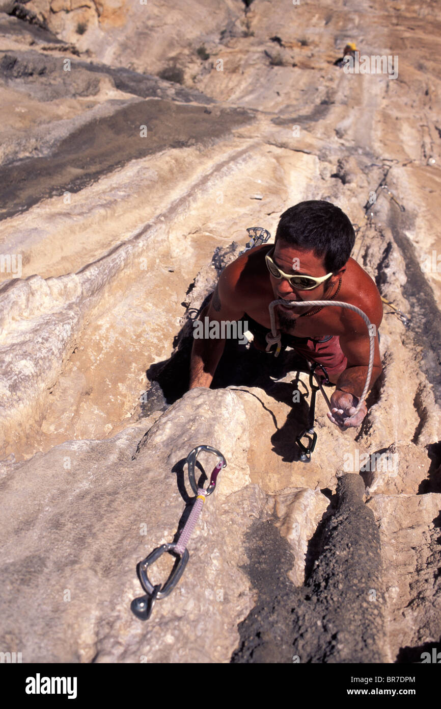 A man biting the rope while climbing a big wall with another man below ...