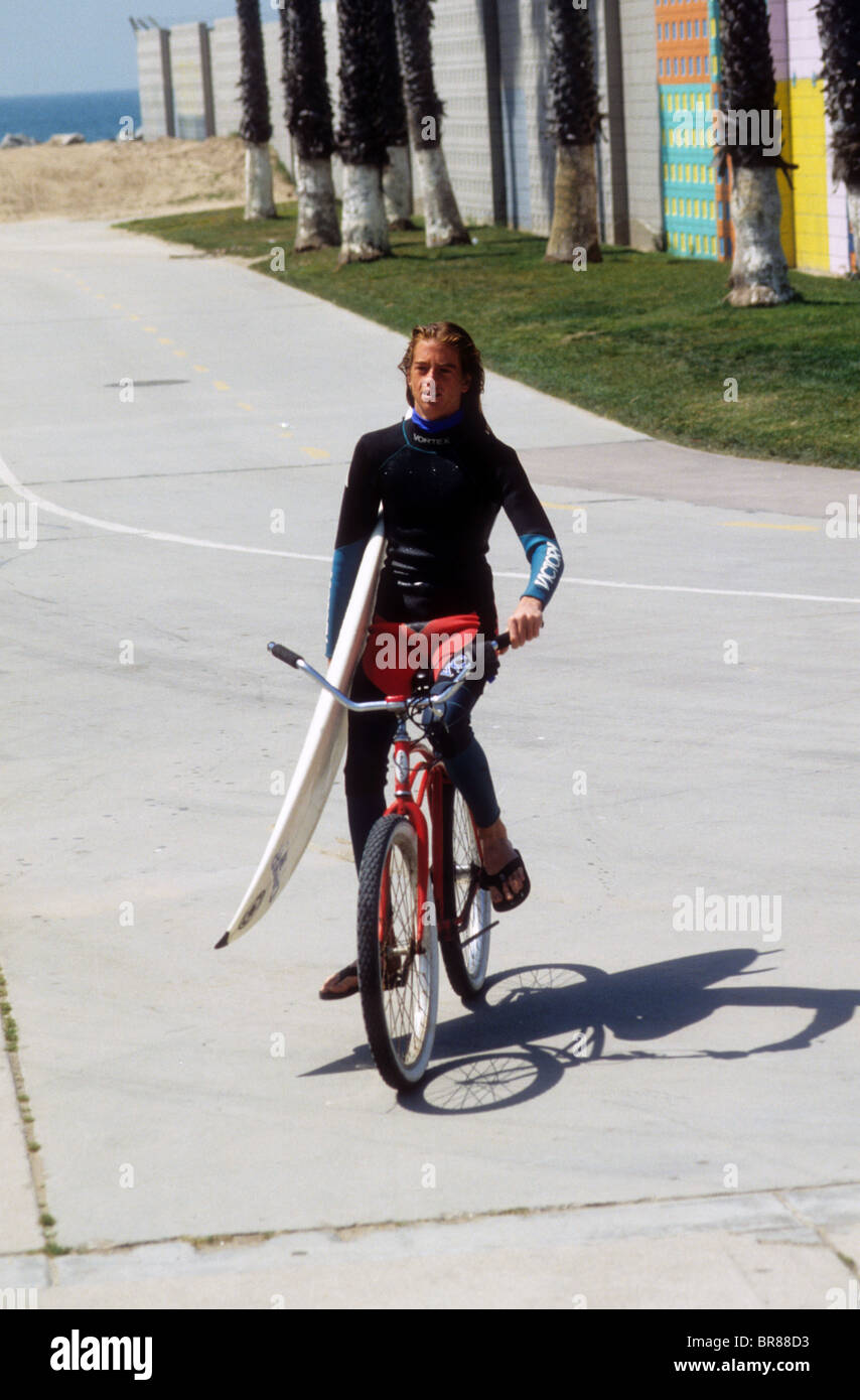 teen male surfer ride bike to beach with board under arm wet suit sun ...